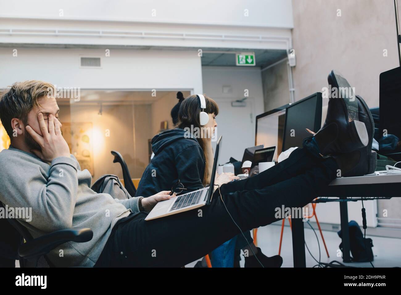 Tired computer programmer sitting with feet up on desk while colleagues ...