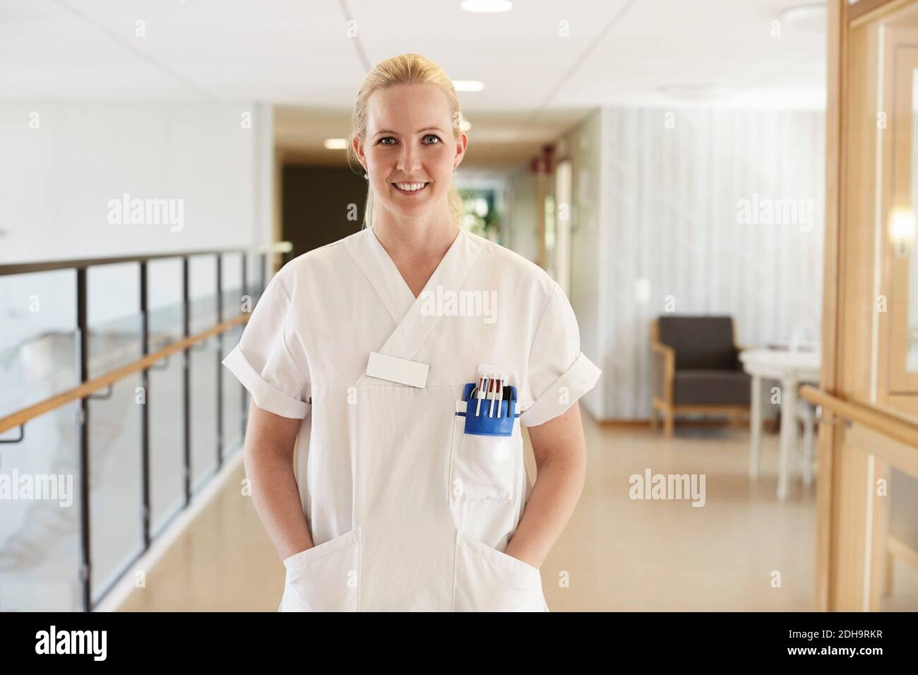 Portrait of happy female nurse standing with hands in pockets at ...