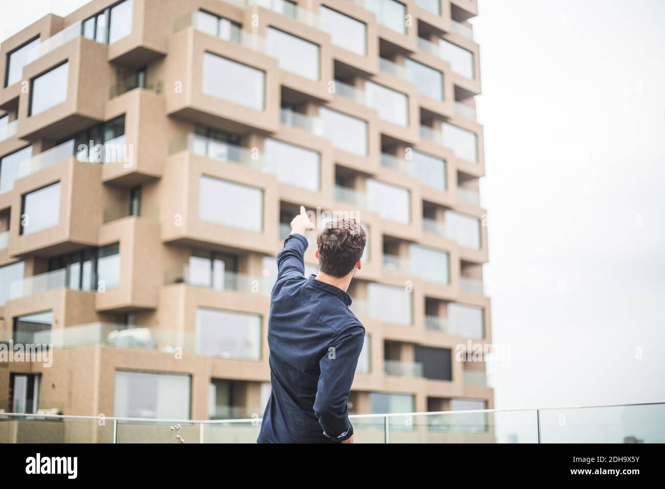 Rear view of man pointing at building while standing by glass railing ...
