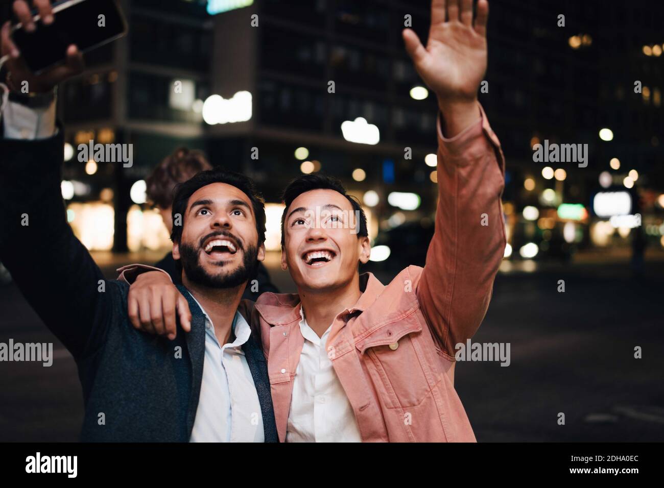 Happy man and male friend looking up while gesturing in city at night ...