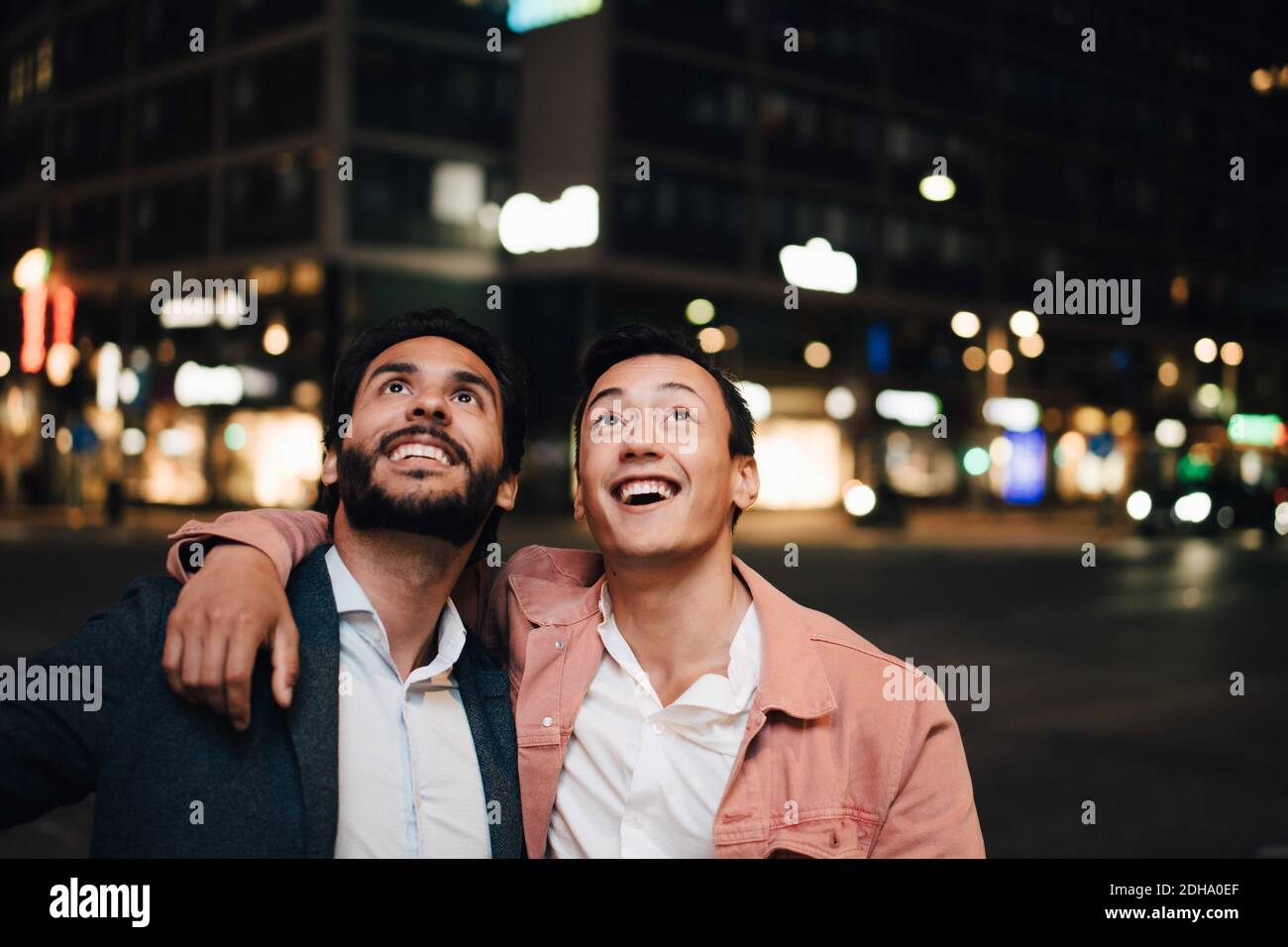Happy man and male friend looking up while standing in city at night ...