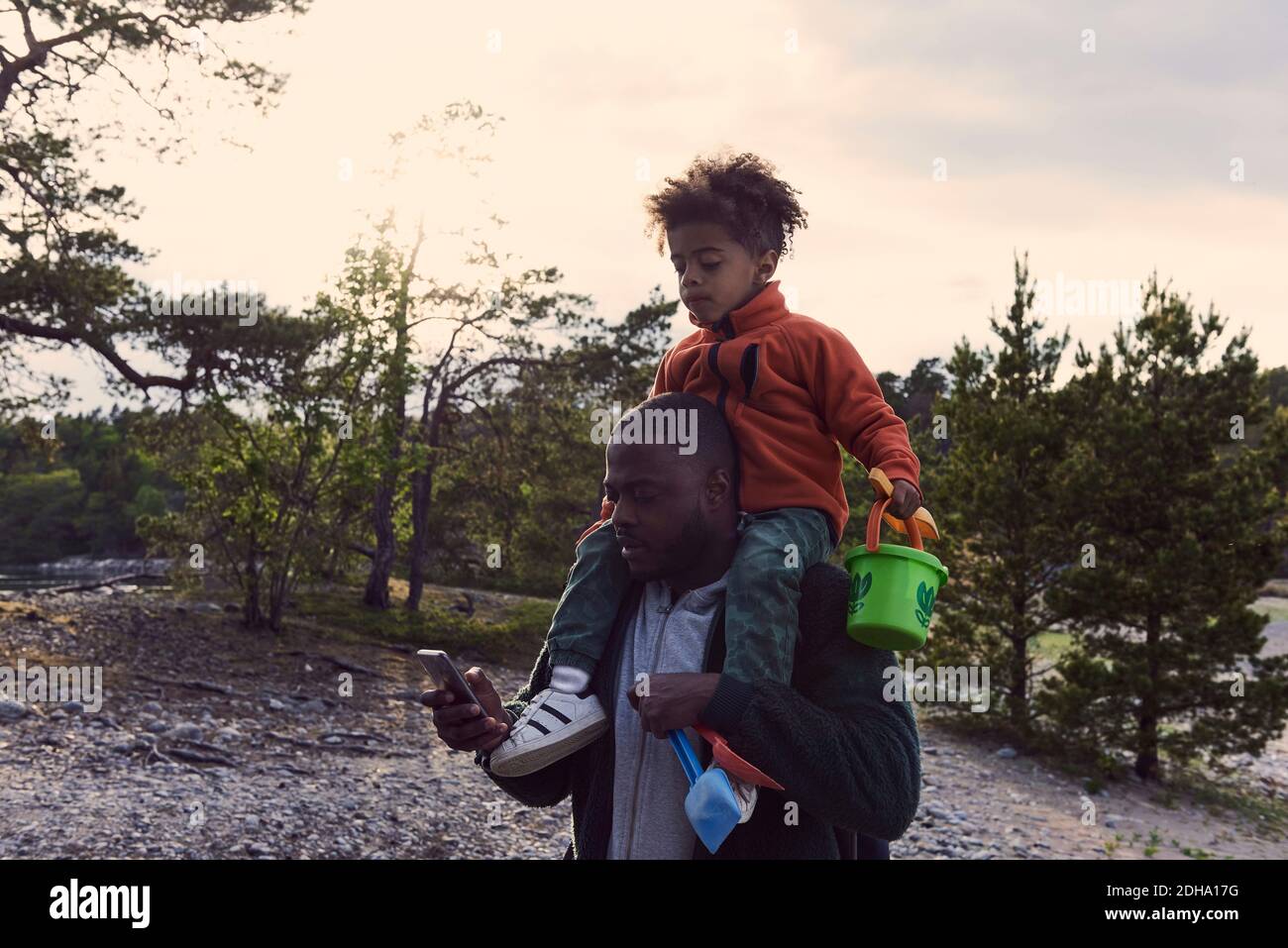 Father using mobile phone while carrying son on shoulders against sky ...