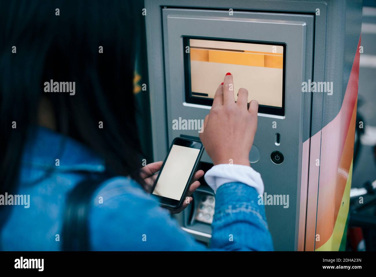Rear view of teenage girl touching ATM screen while holding smart phone ...