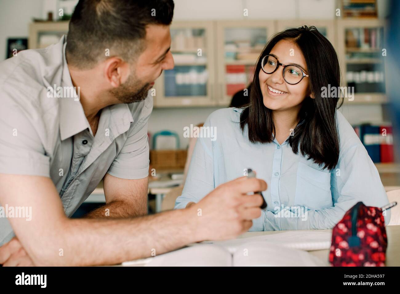 Smiling male teacher explaining student while leaning on table in ...