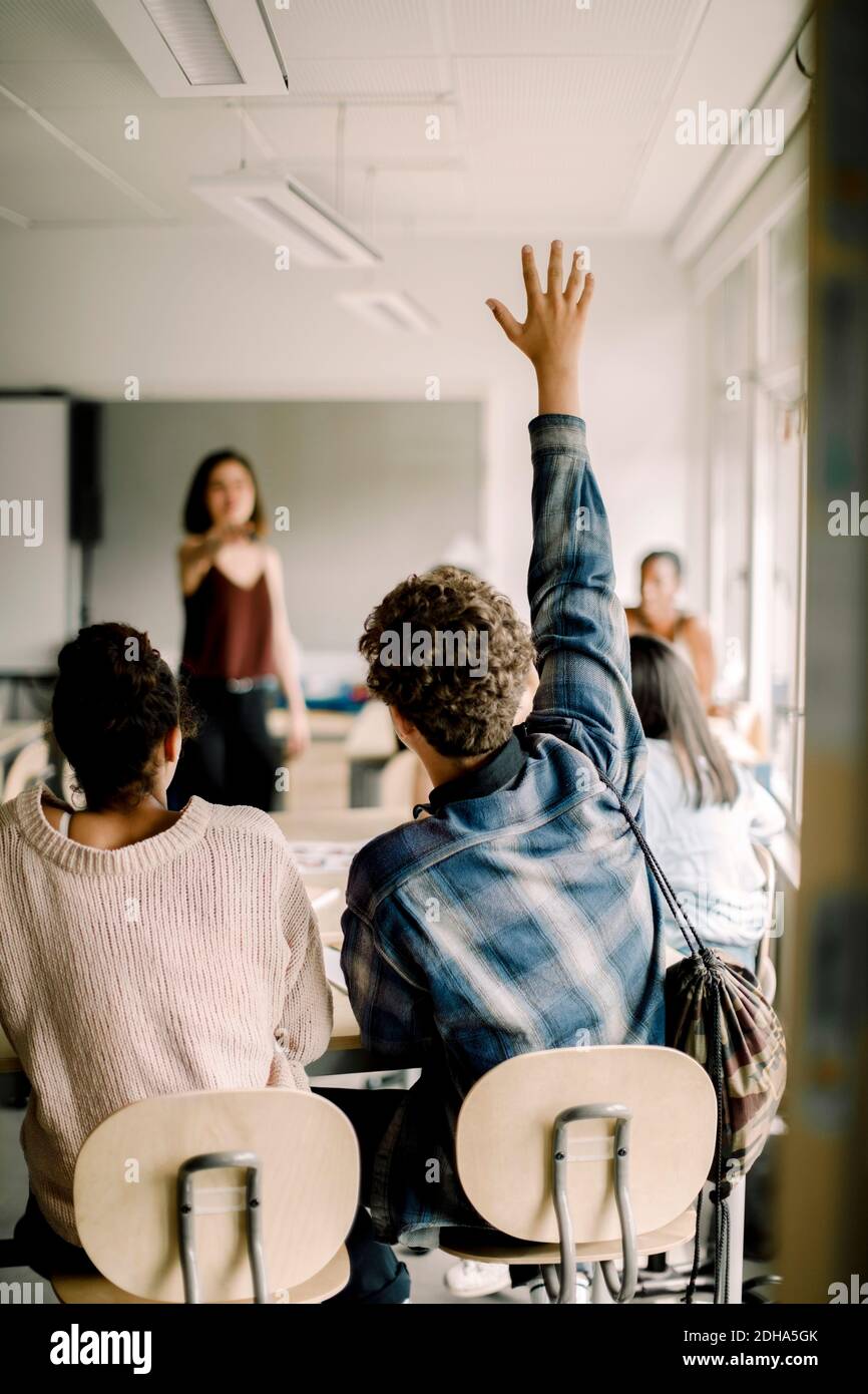 Male student with hand raised while female teacher pointing in ...