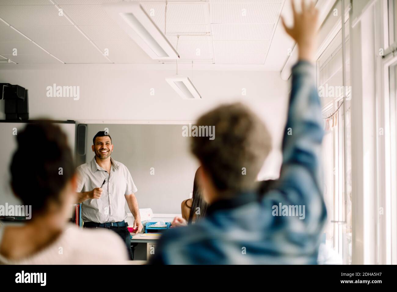 Male student with hand raised while smiling tutor standing in classroom ...