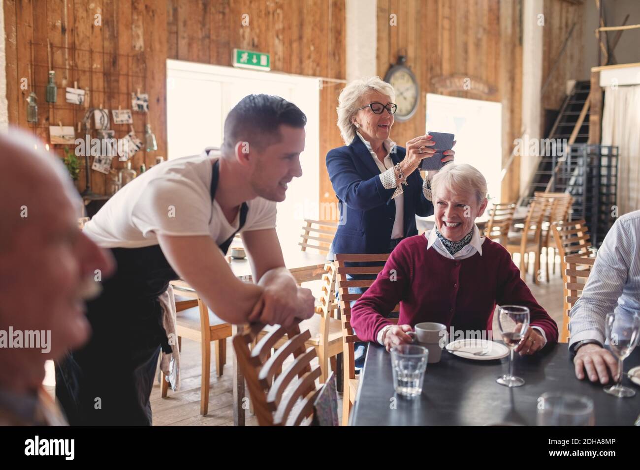 Young waiter leaning on chair while senior friends socializing in ...