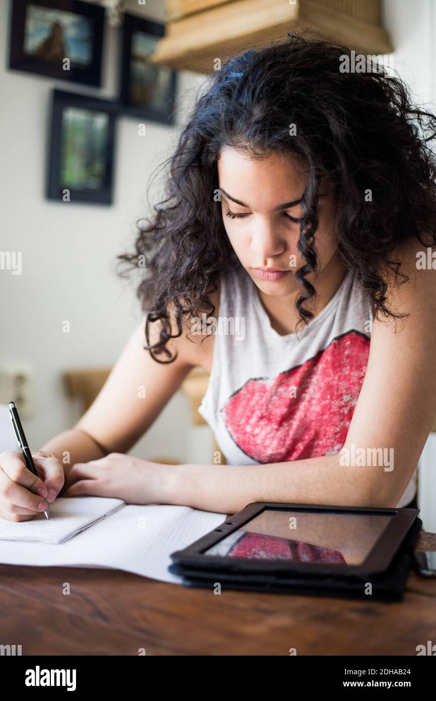 Teenage girl studying at table while writing in book by digital tablet ...