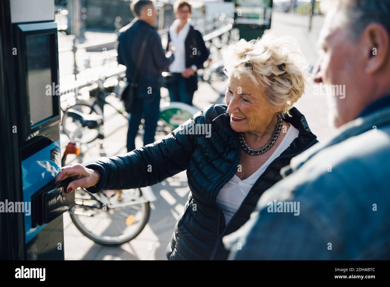 Senior woman operating bike vending machine by man on sidewalk in city ...