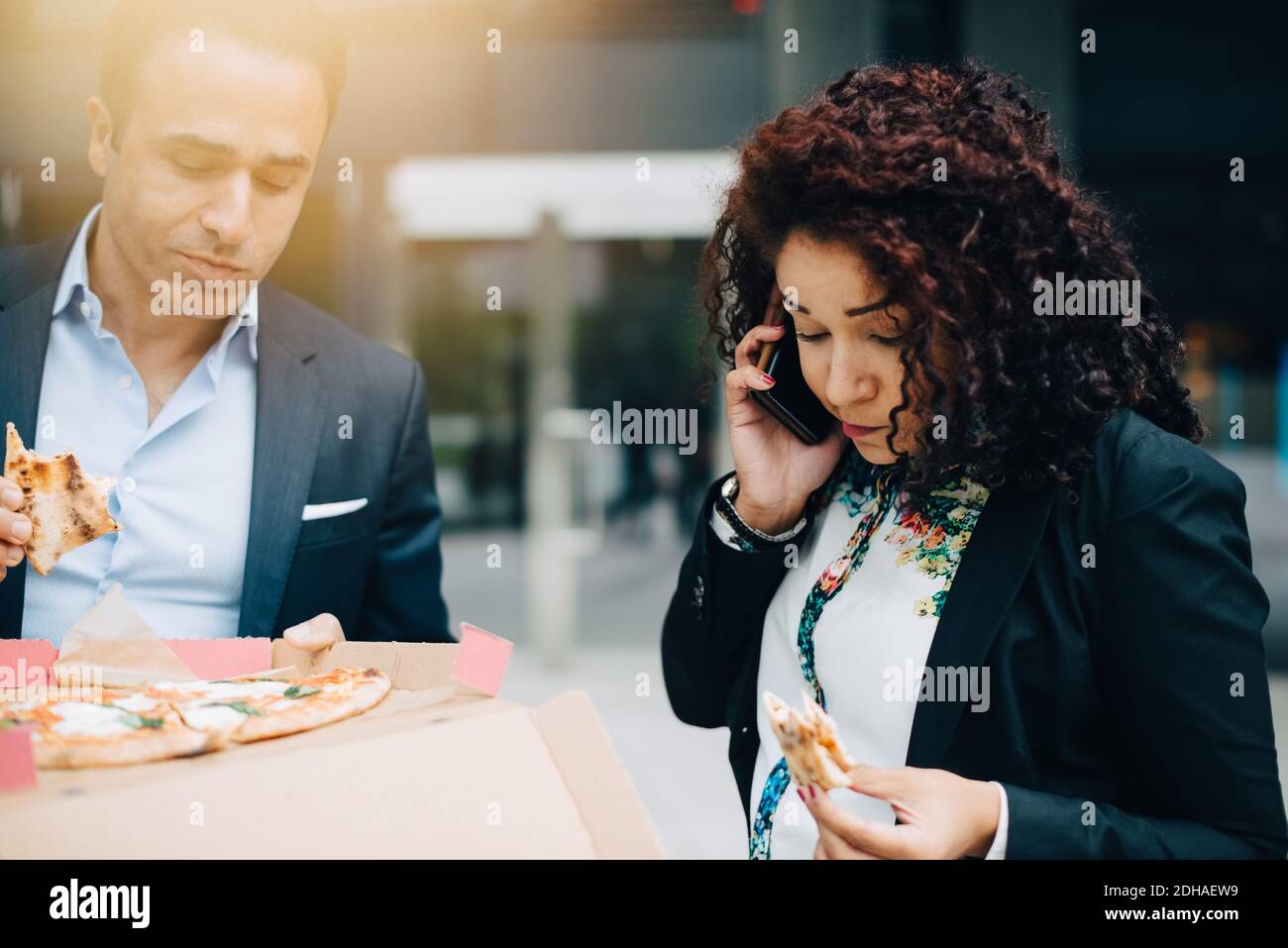 Male and female business colleagues eating pizza against building ...
