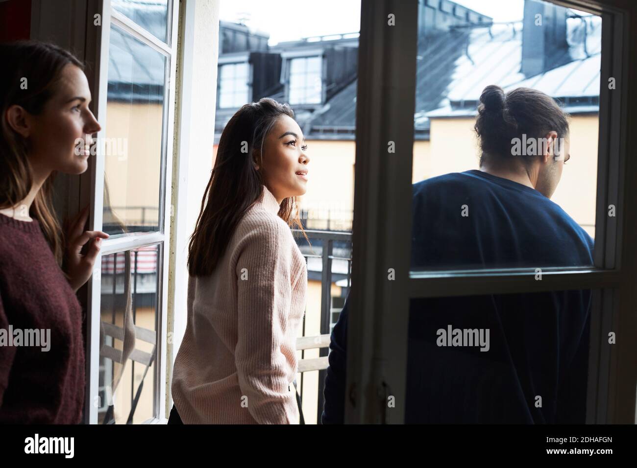 Smiling young friends looking through window while standing in balcony ...