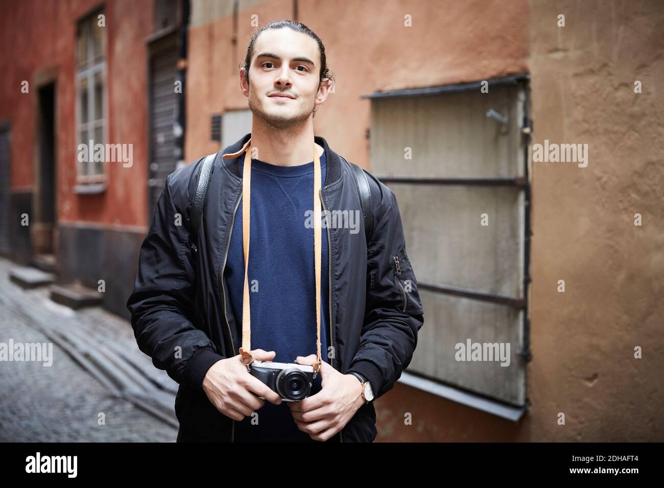 Portrait of young man holding camera while standing by building in ...