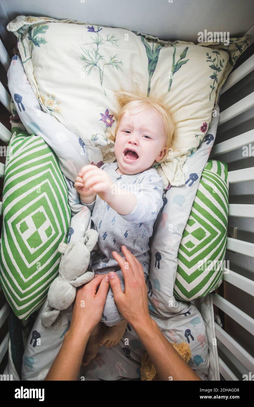 Crying baby boy lying in crib while hands of father reaching for him ...