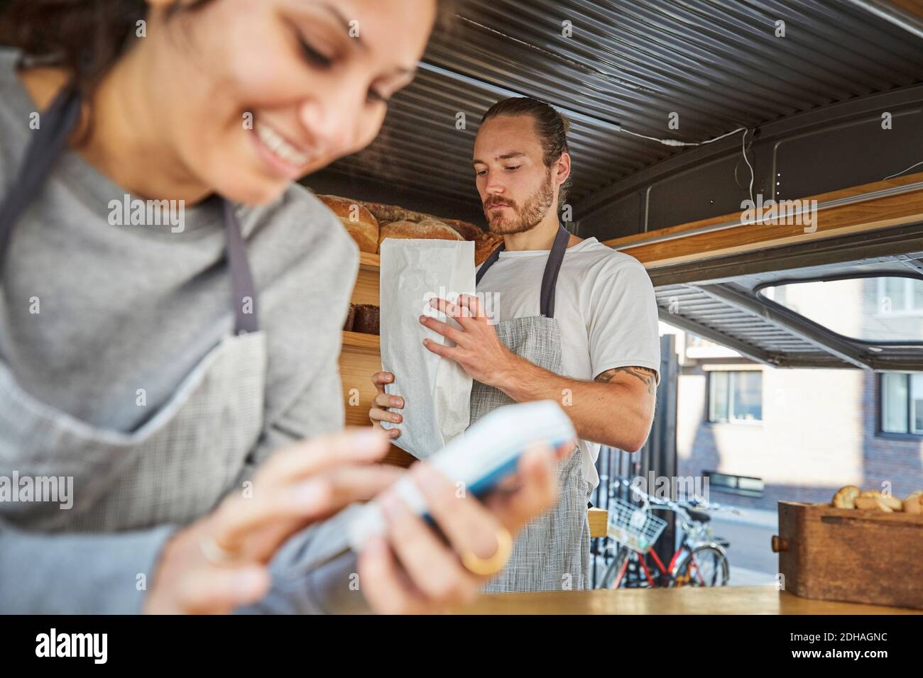 Male owner packing bread in paper bag while colleague using credit card ...