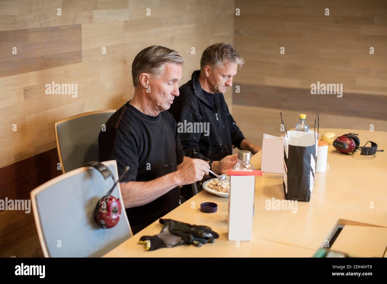 Workers having lunch at table while sitting against wall in industry ...