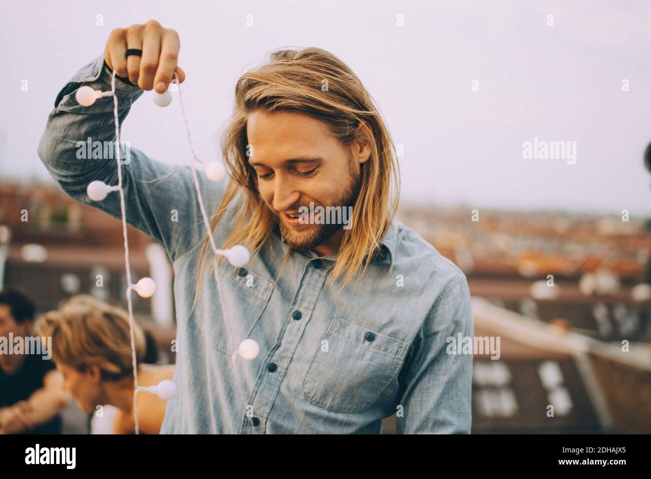 Smiling young man with string light standing on terrace during party ...