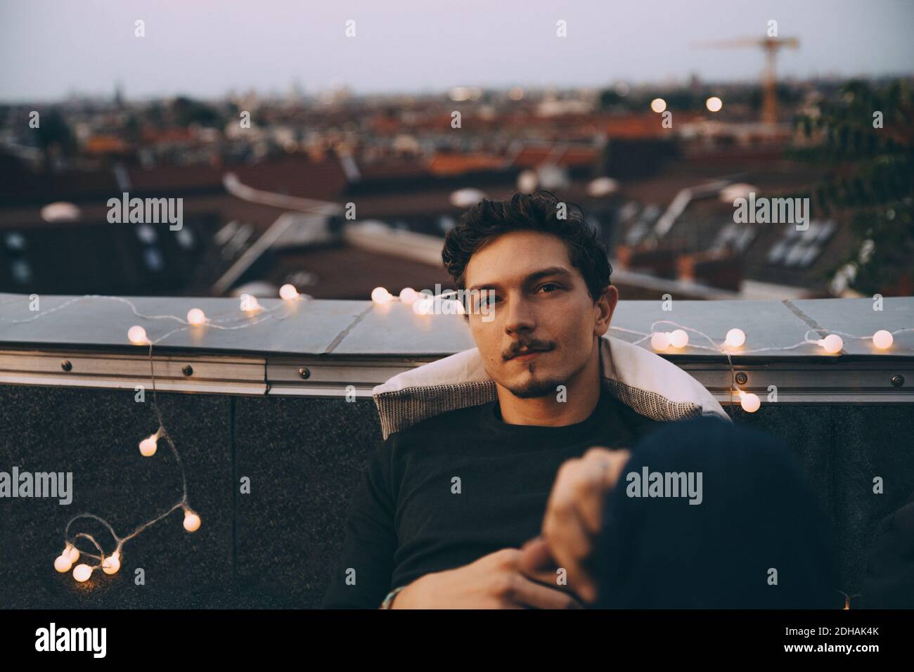 Portrait of young man sitting on illuminated terrace in city during ...