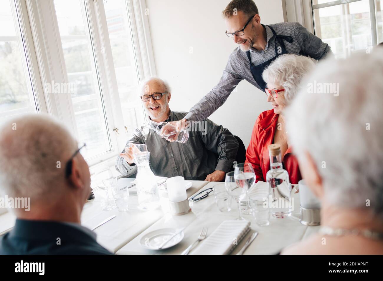 Owner serving water to smiling senior man sitting with friends in ...
