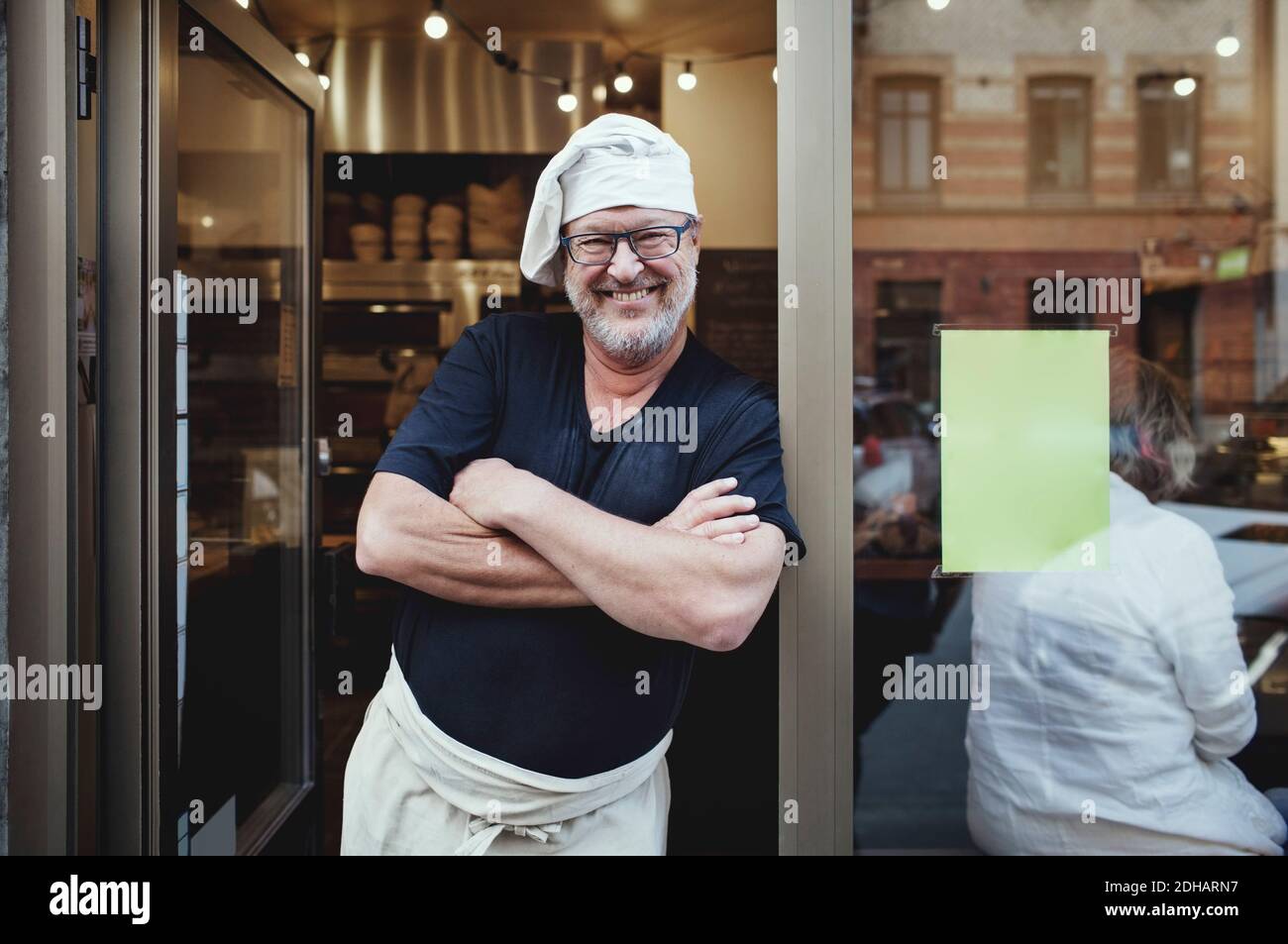 Smiling male baker standing with arms crossed at entrance of bakery ...