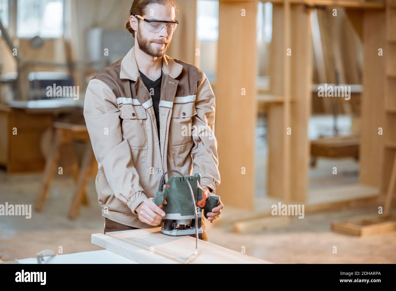 Handsome carpenter in uniform chamfers wooden bar with a hand machine ...