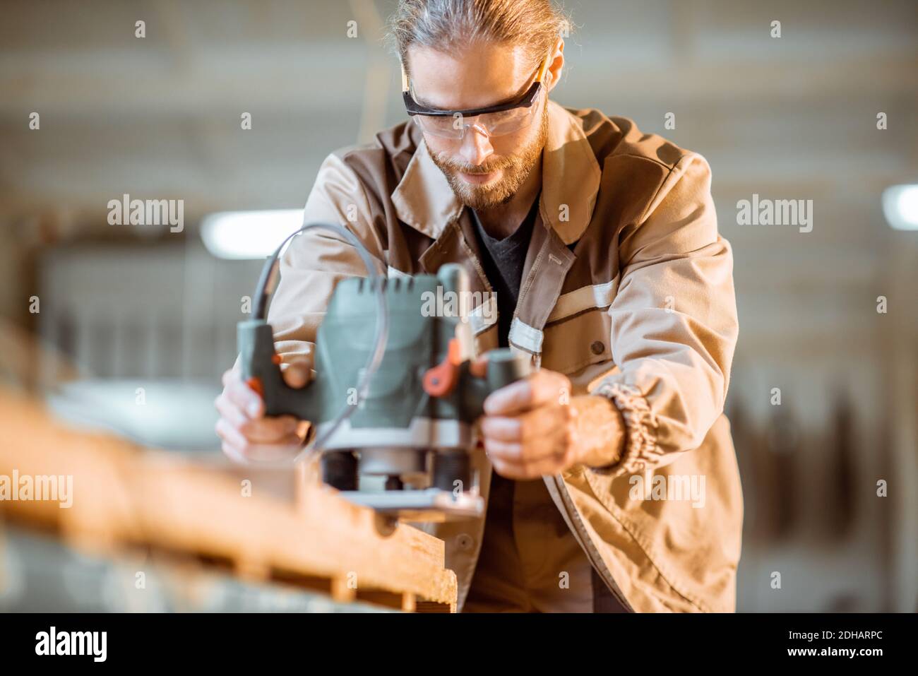 Handsome carpenter in uniform chamfers wooden bar with a hand machine ...