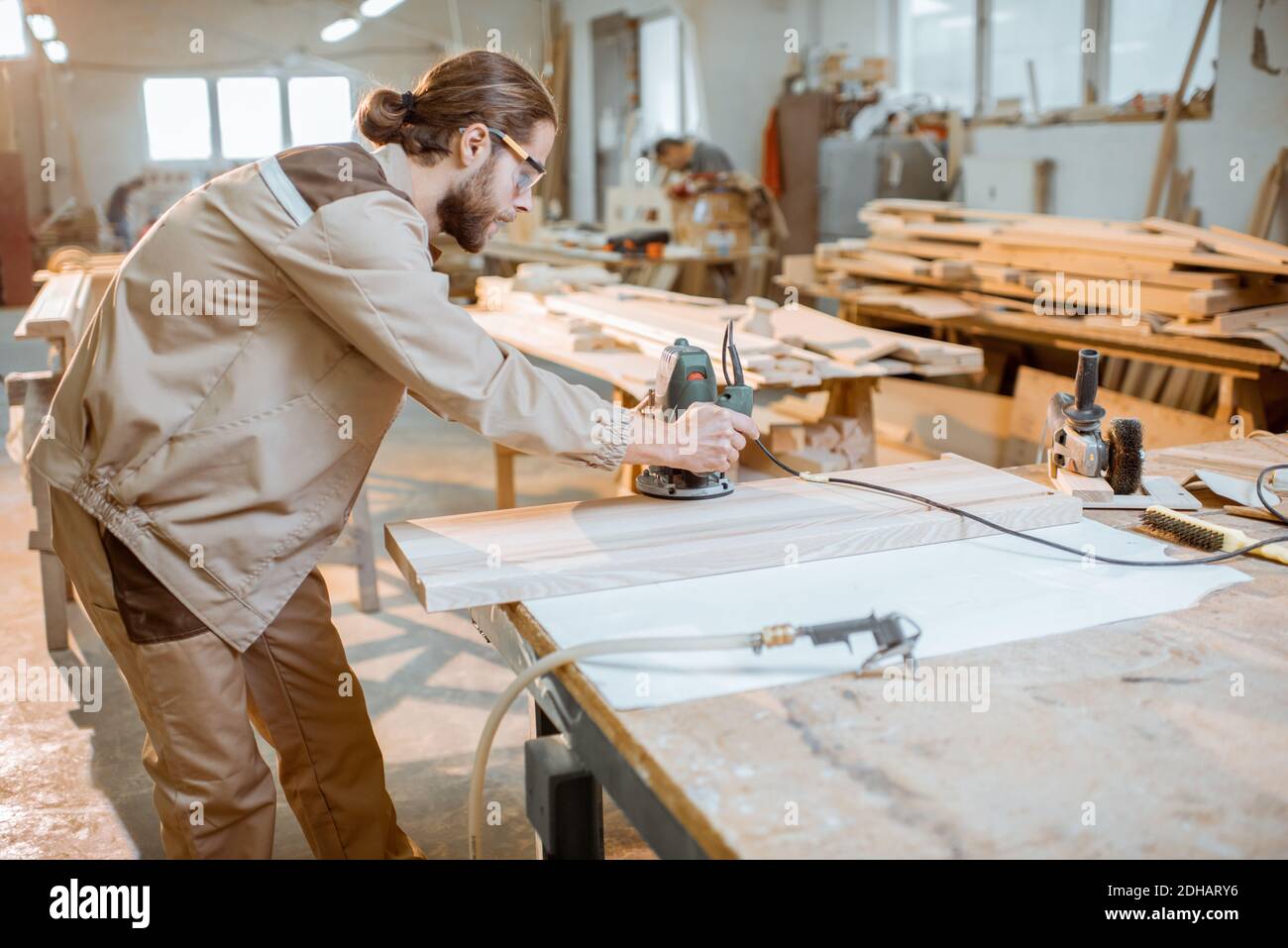 Handsome carpenter in uniform chamfers wooden bar with a hand machine ...