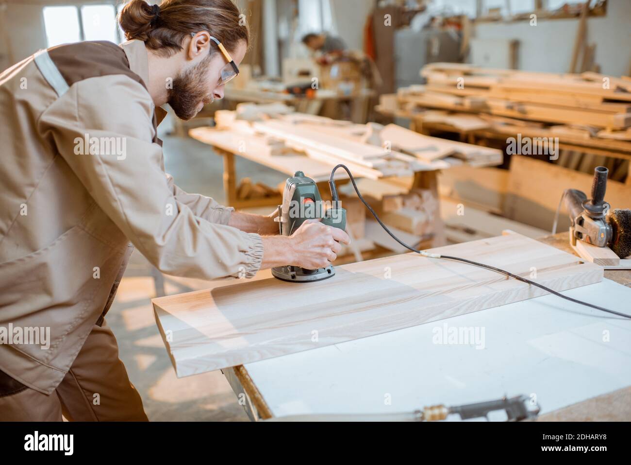 Handsome carpenter in uniform chamfers wooden bar with a hand machine ...