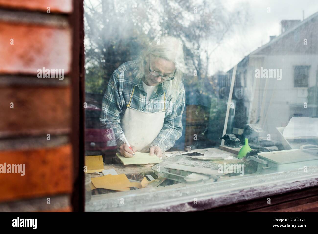 Senior craftsperson writing on paper seen through glass window Stock ...