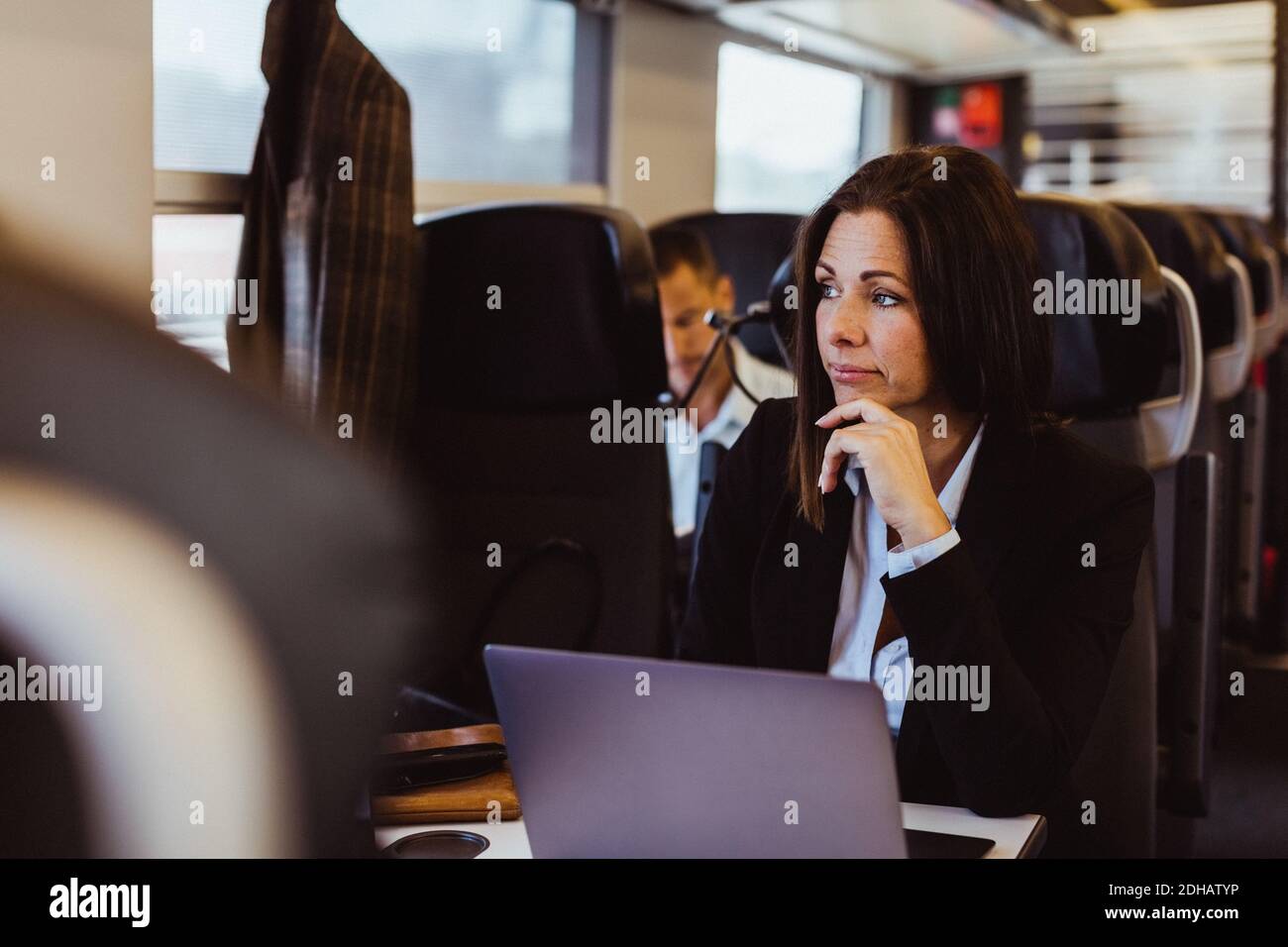 Thoughtful business commuter using laptop while looking away in train ...