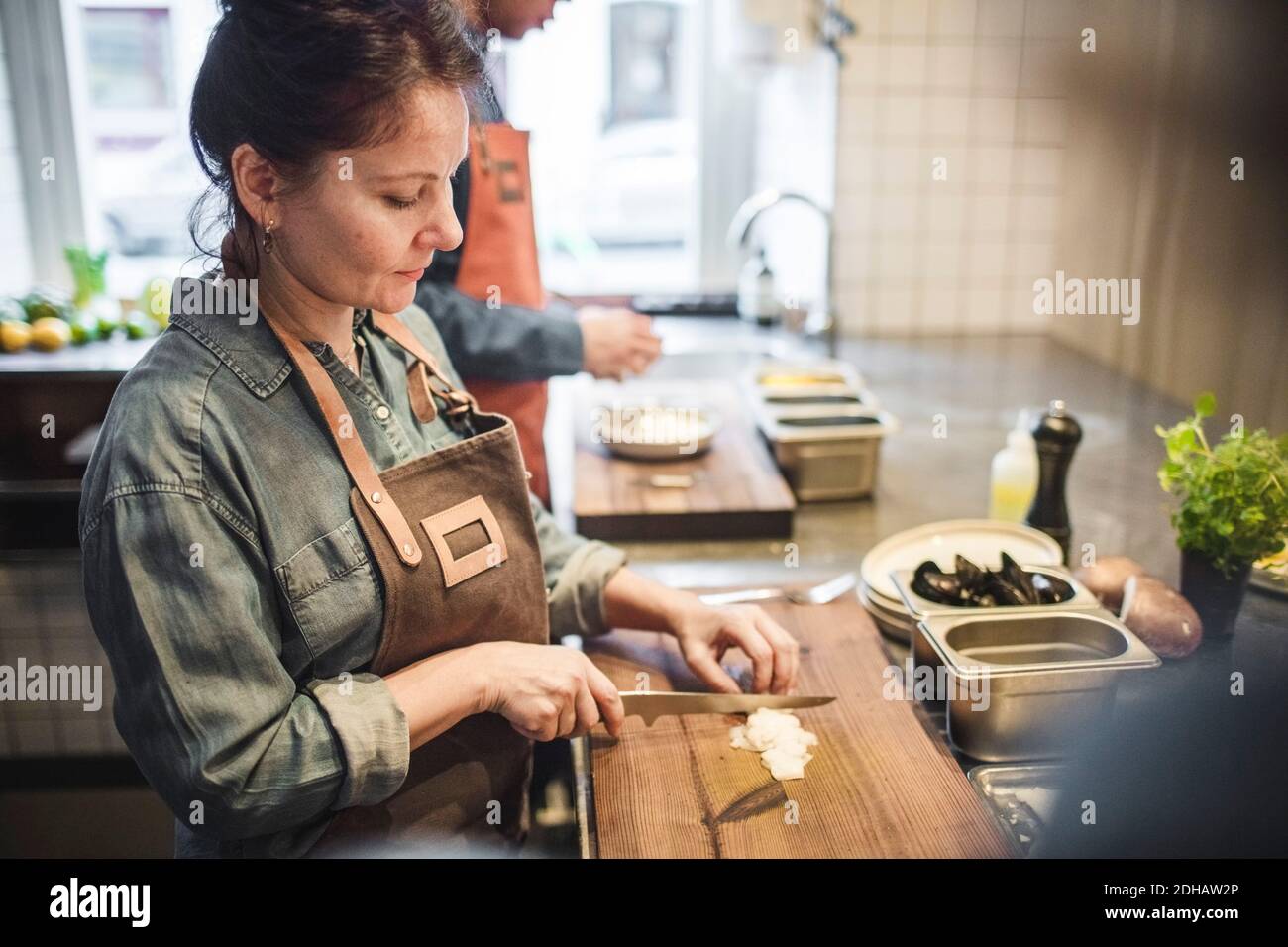 Mature female chef cutting food on board at counter in restaurant ...
