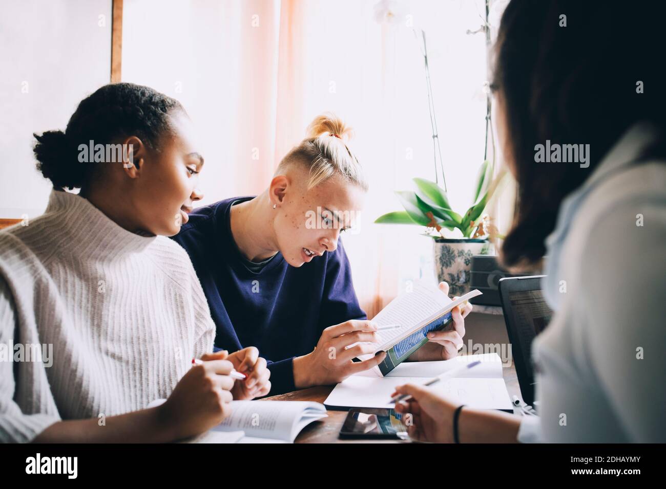 Teenage students discussing over book while sitting together at table ...