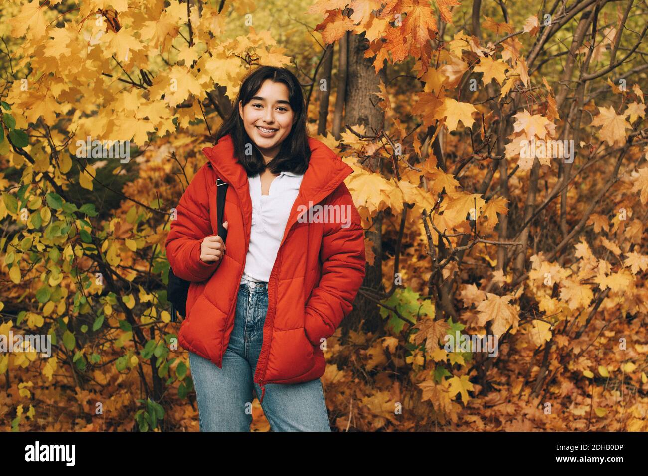 Portrait of smiling teenage girl standing with backpack against autumn ...