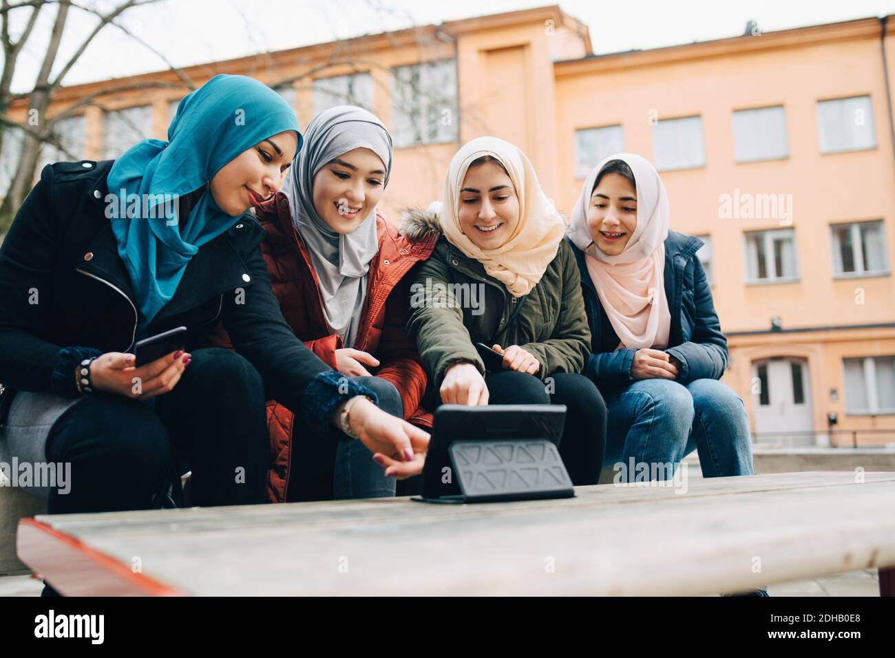 Happy female Muslim friends sitting with digital tablet on bench in ...