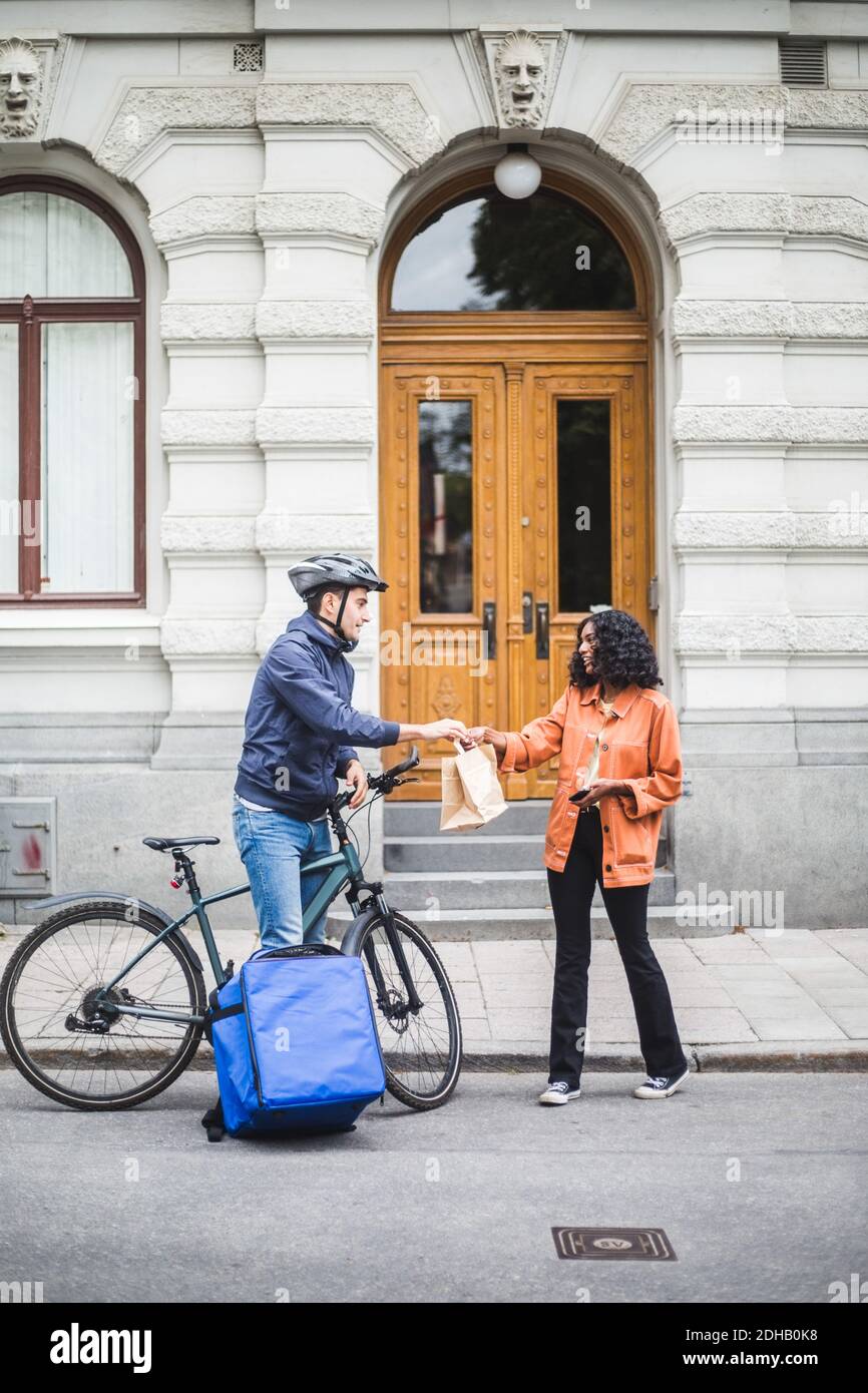 Food delivery man delivering package to woman standing on sidewalk in ...