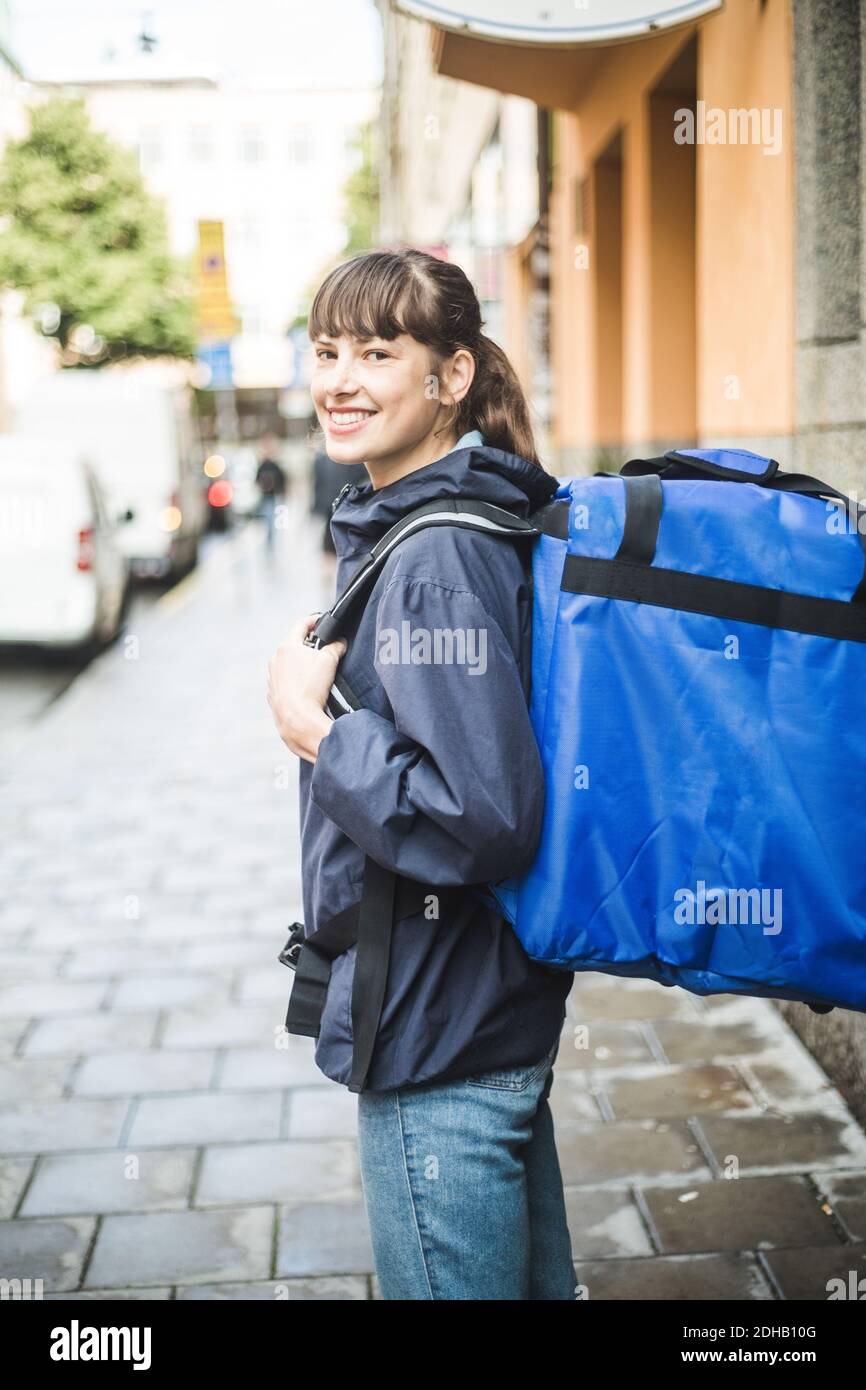Portrait of confident delivery woman with bag standing on sidewalk ...