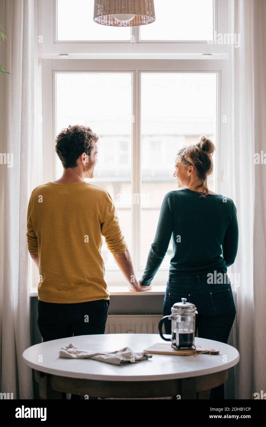 Rear view of romantic couple standing at window in living room Stock ...