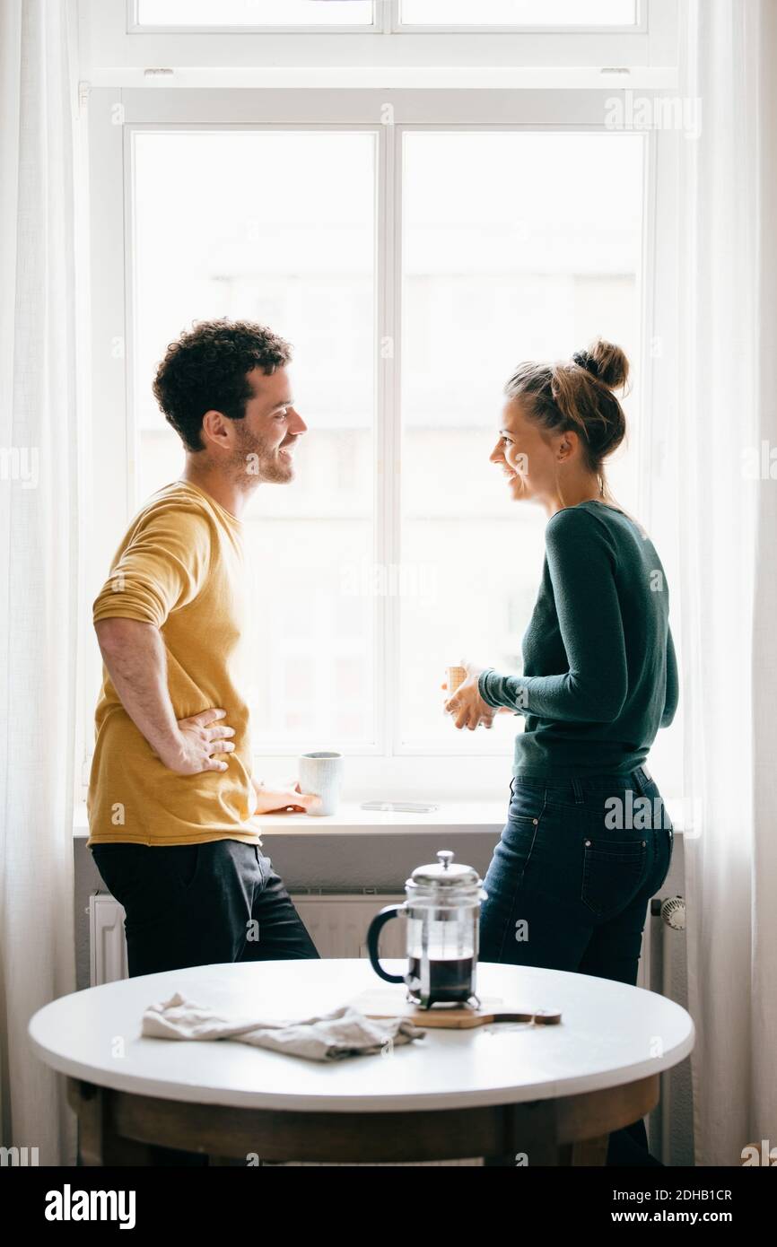 Side view of happy couple talking while standing by window at home ...