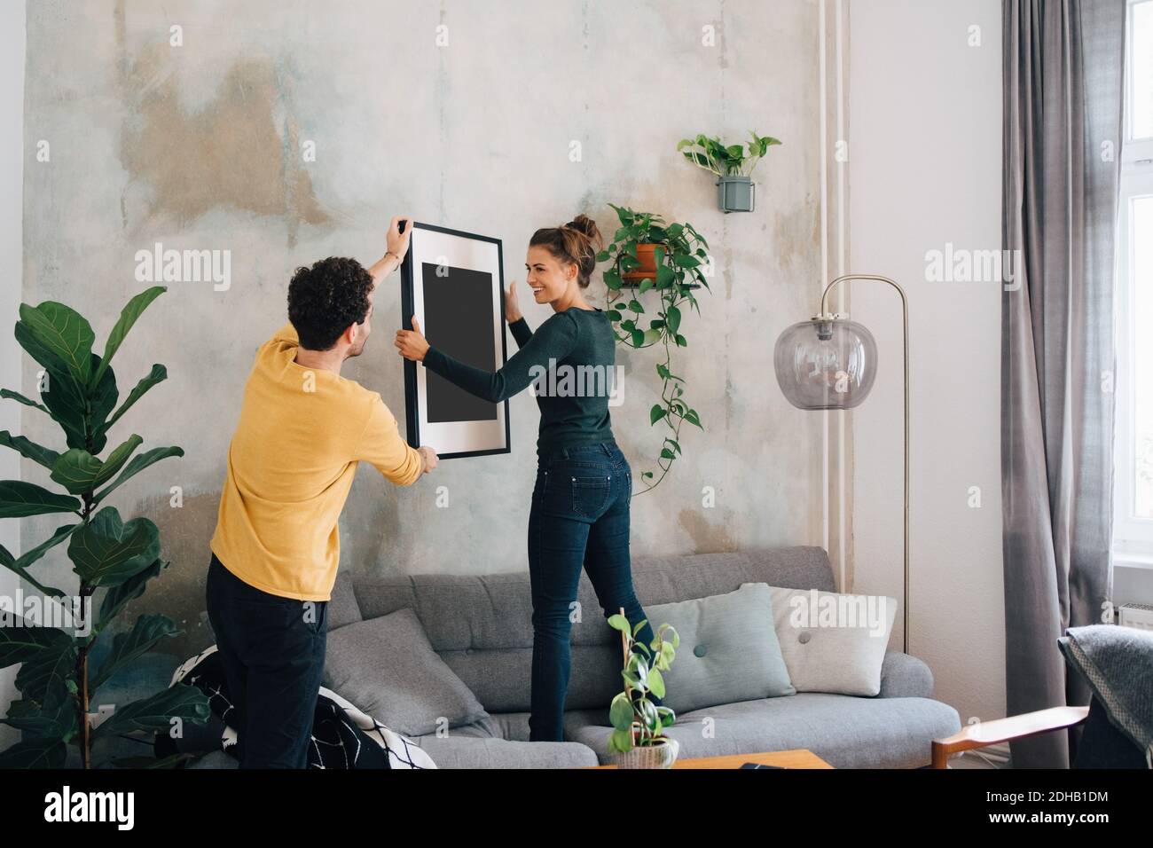 Mid adult man passing picture frame to girlfriend against wall at new ...
