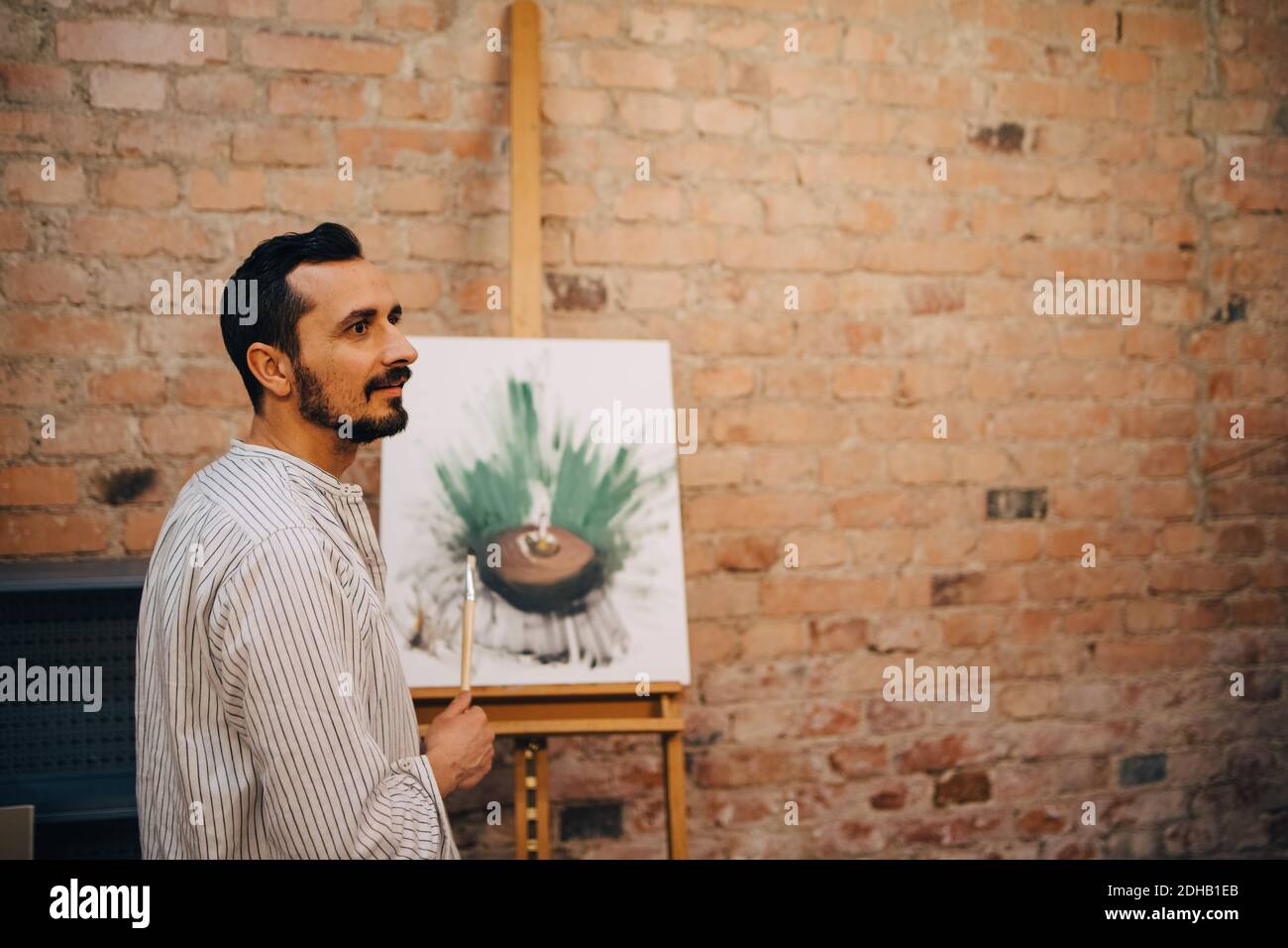 Side view of male artist standing by painting against brick wall at ...