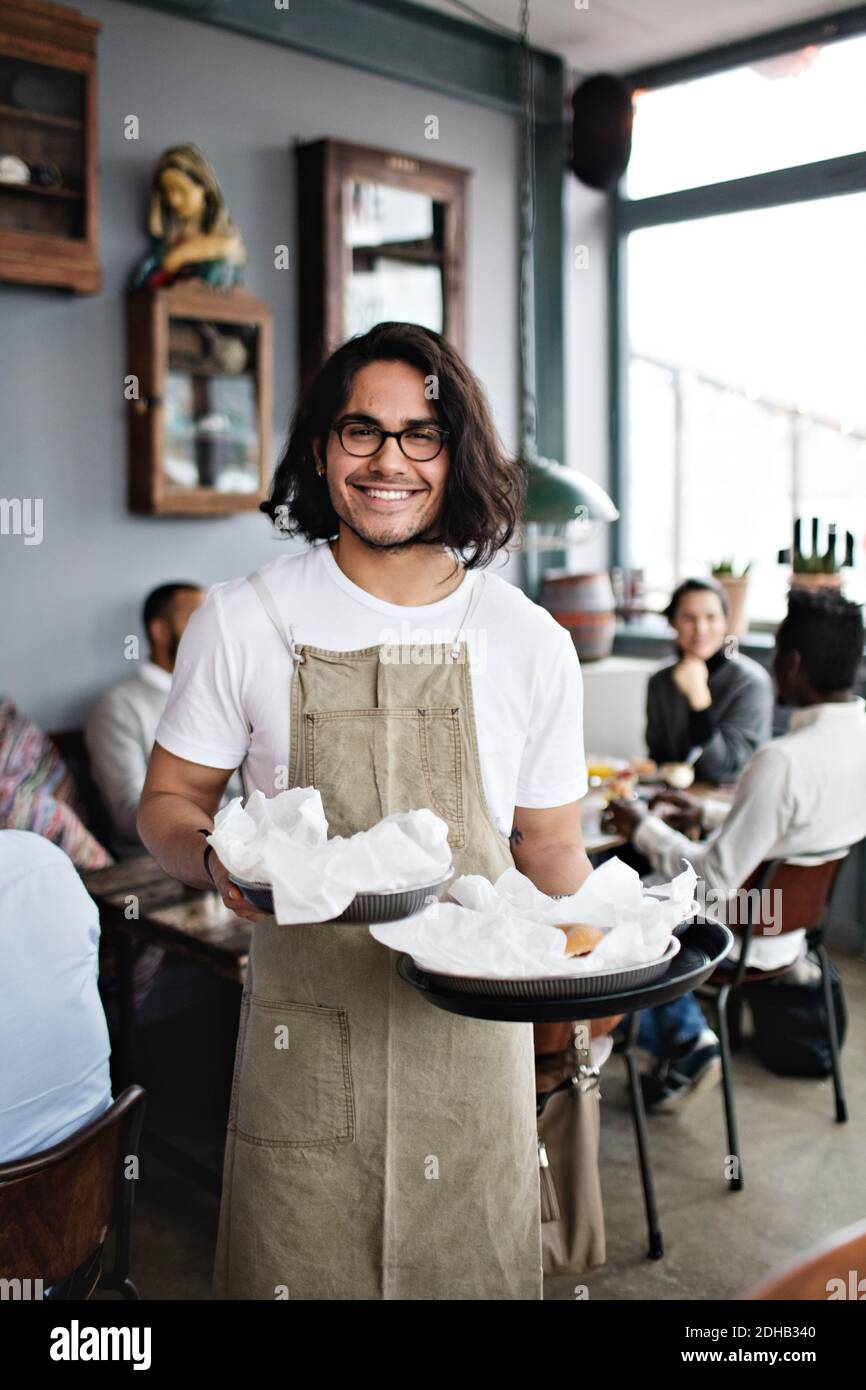 Portrait of confident young waiter serving food while standing against ...