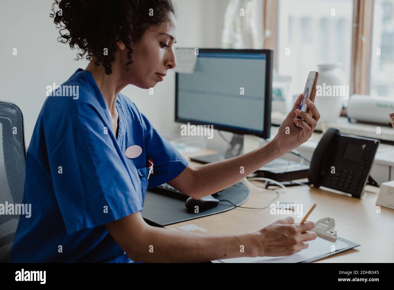 Nurse writing in clipboard while using phone for video conference at ...
