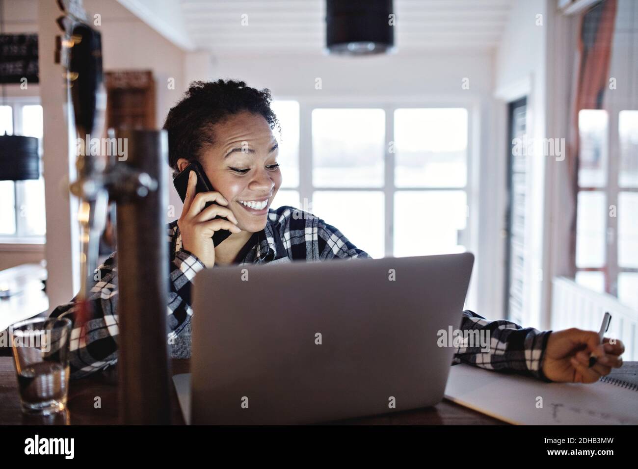 Happy female manager talking on mobile phone while using laptop at bar ...