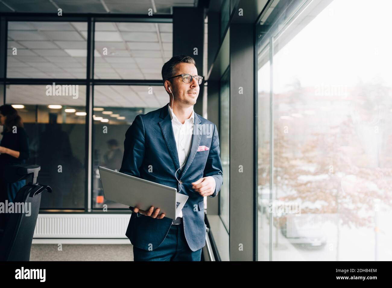 Smiling entrepreneur holding laptop while looking through window in ...