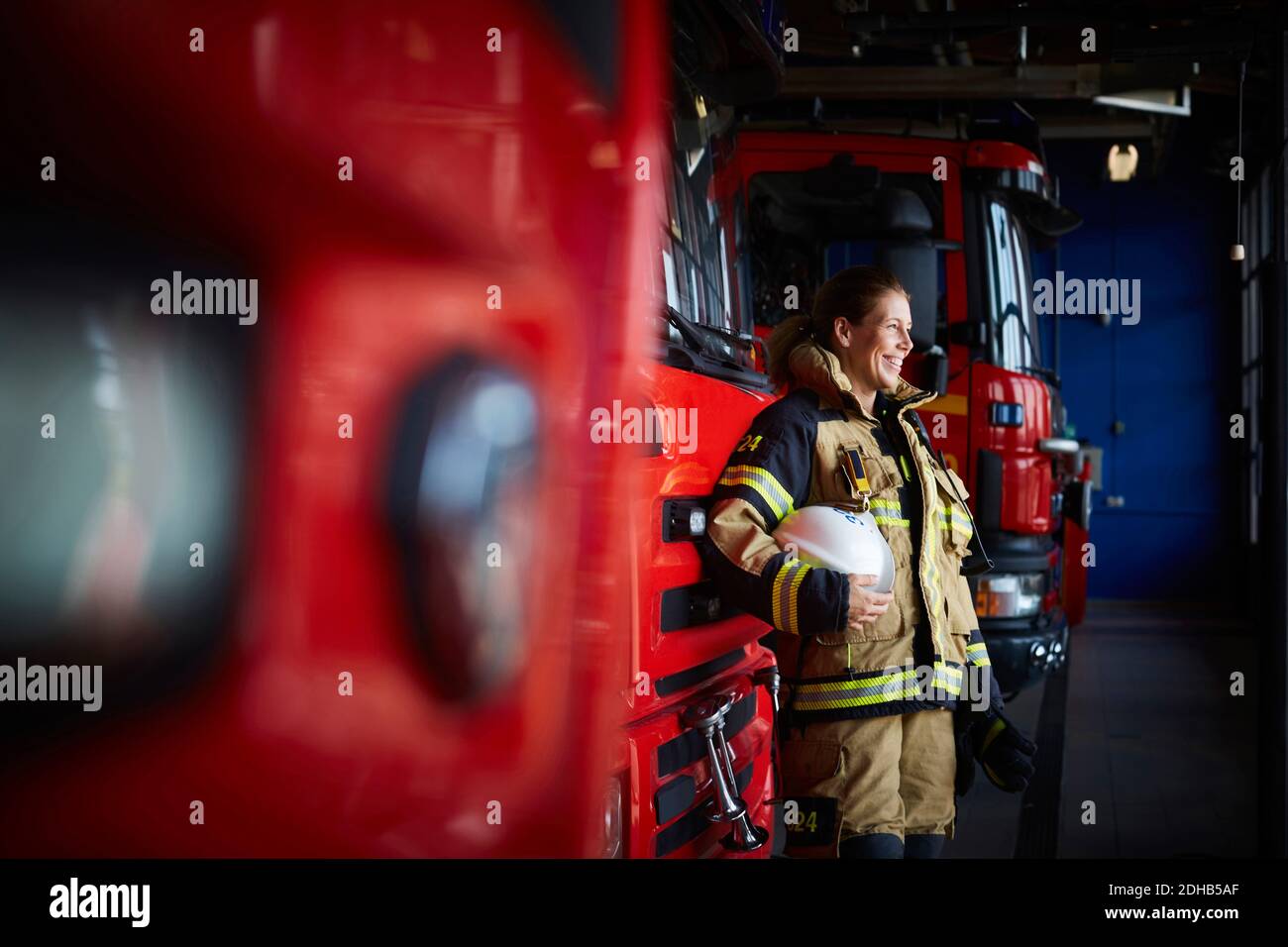 Smiling female firefighter standing by fire engine at fire station ...