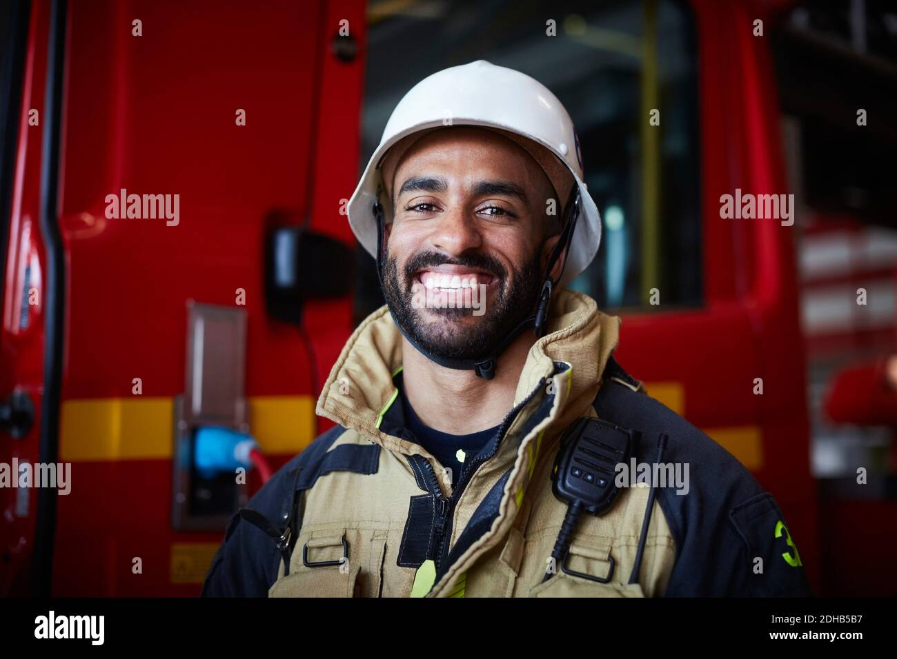 Portrait of smiling firefighter wearing helmet standing at fire station ...