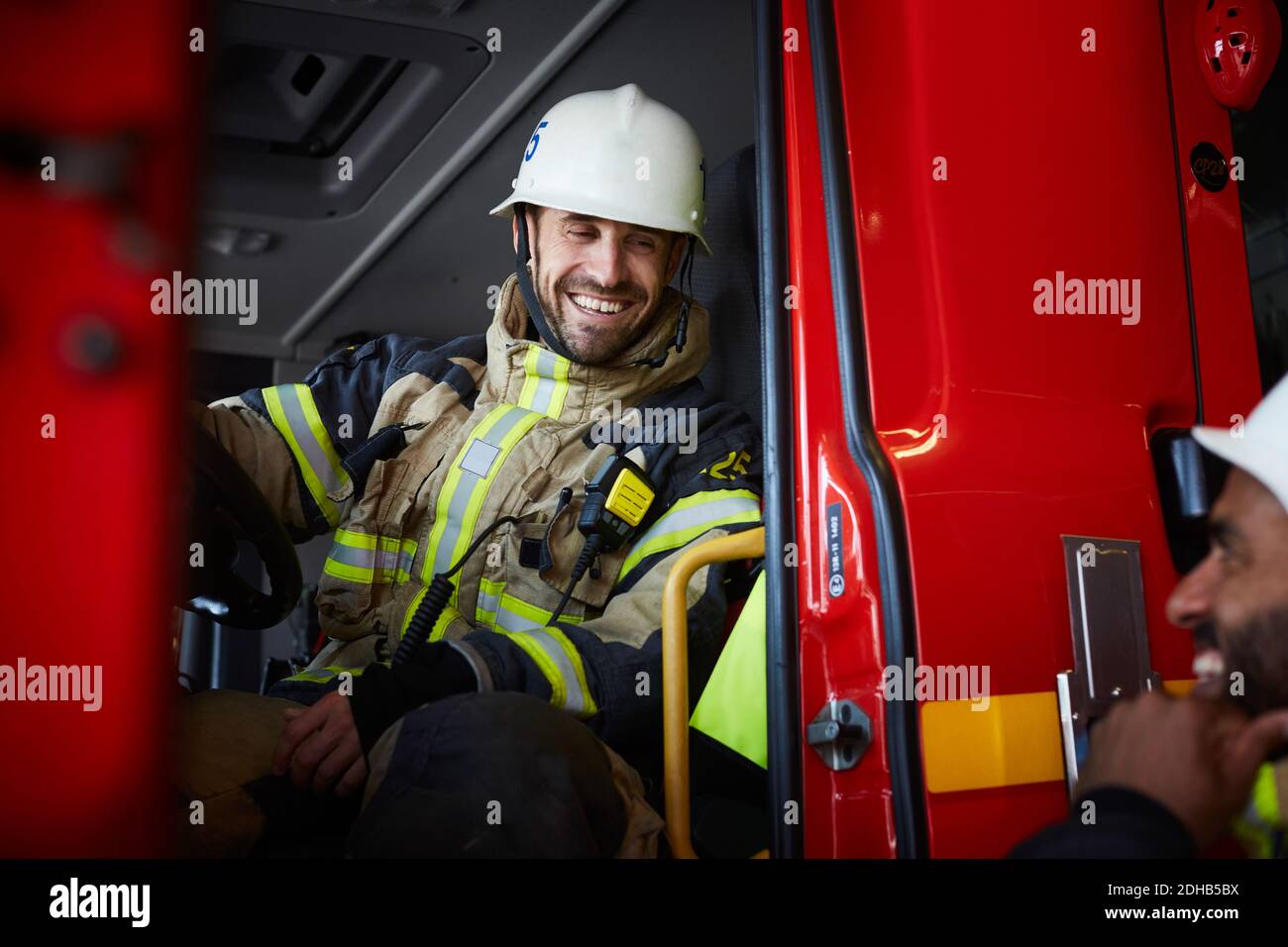 Smiling firefighter sitting in fire engine while talking to coworker ...