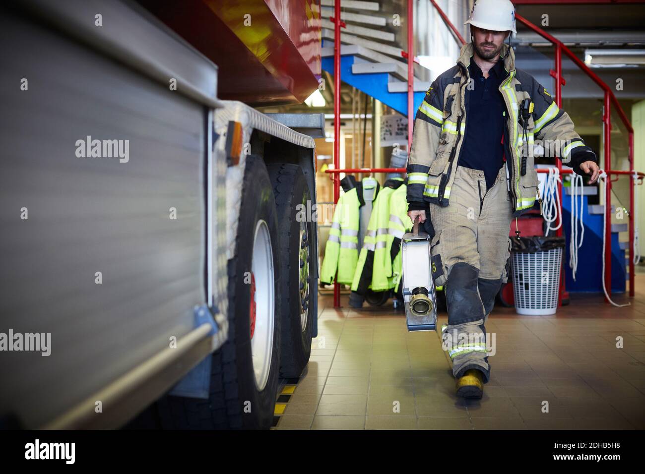 Full length of firefighter holding fire hose while walking by fire ...
