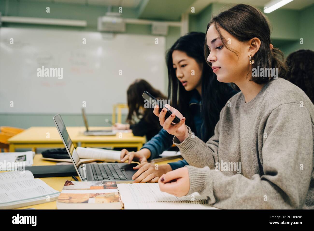 Female high school student using smart phone while sitting by classmate ...