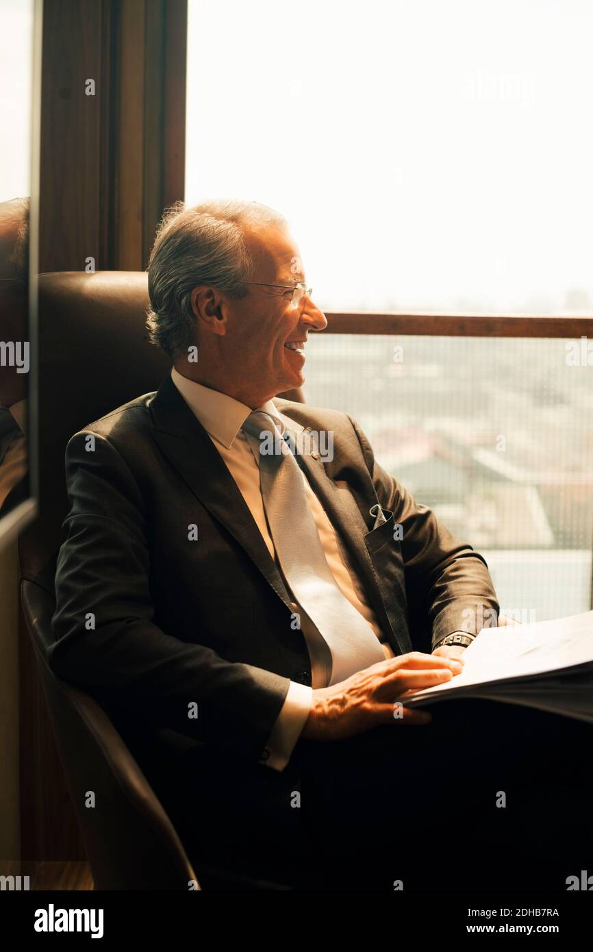 Smiling mature lawyer with documents looking through window at office ...