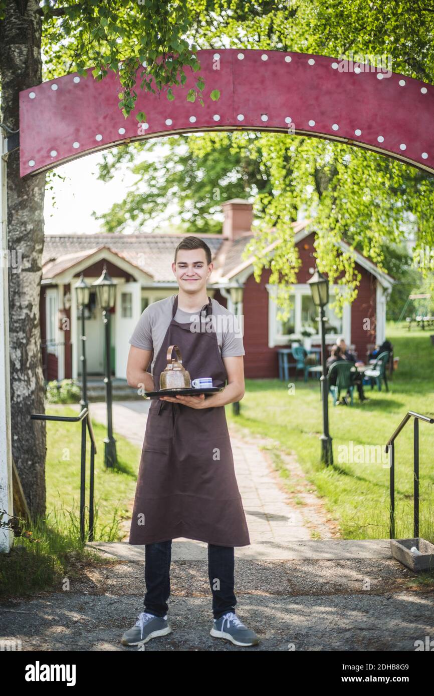 Full length portrait of smiling young waiter holding serving tray while ...